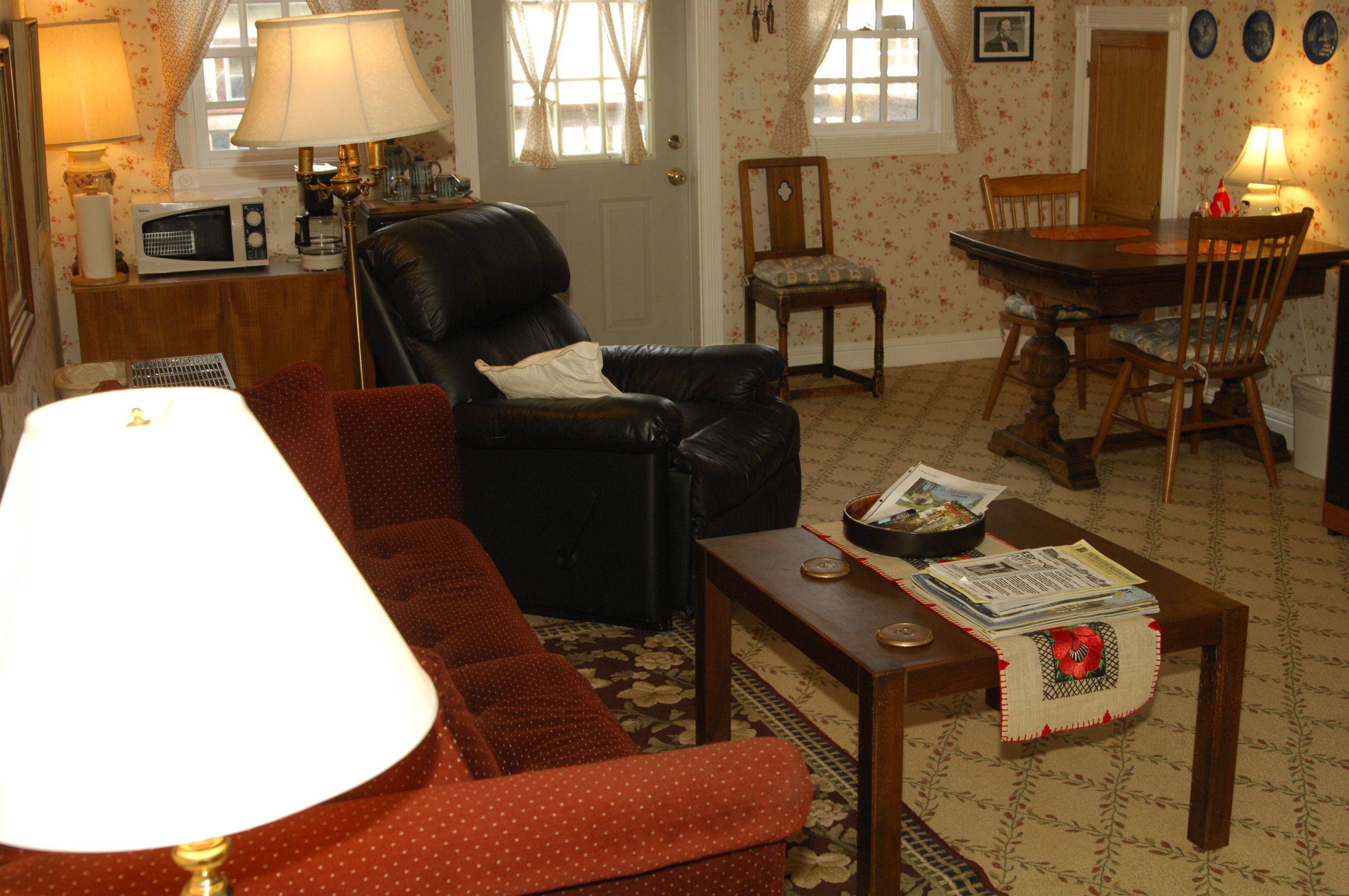 Uma sala de estar com um sofá, uma poltrona, uma mesa de centro e uma mesa de jantar com cadeiras. Há uma luminária na mesa e um micro-ondas numa bancada.