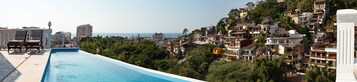 Piscina infinita no terraço, com espreguiçadeiras, com vista para uma encosta urbana densa e o mar.