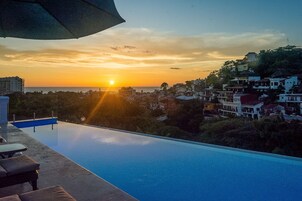 Piscina com vista para o pôr do sol, cadeiras de descanso e um edifício ao fundo.