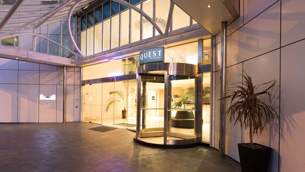 Entrada de hotel com porta giratória, placa com a inscrição 'QUEST em Durham' e um vaso de planta.