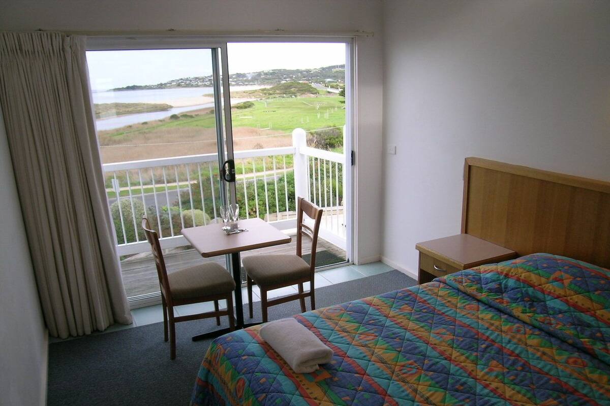 Quarto de hotel com uma cama, uma pequena mesa com cadeiras, uma varanda com vista para a praia e o mar, e uma cabeceira de madeira.