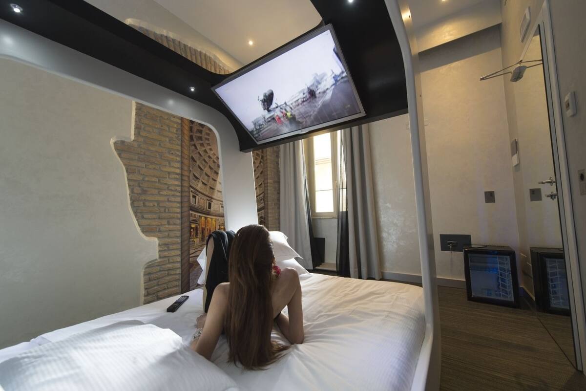Quarto de hotel com cama, televisão fixada na parede, janela com cortinas e design de parede de tijolos.