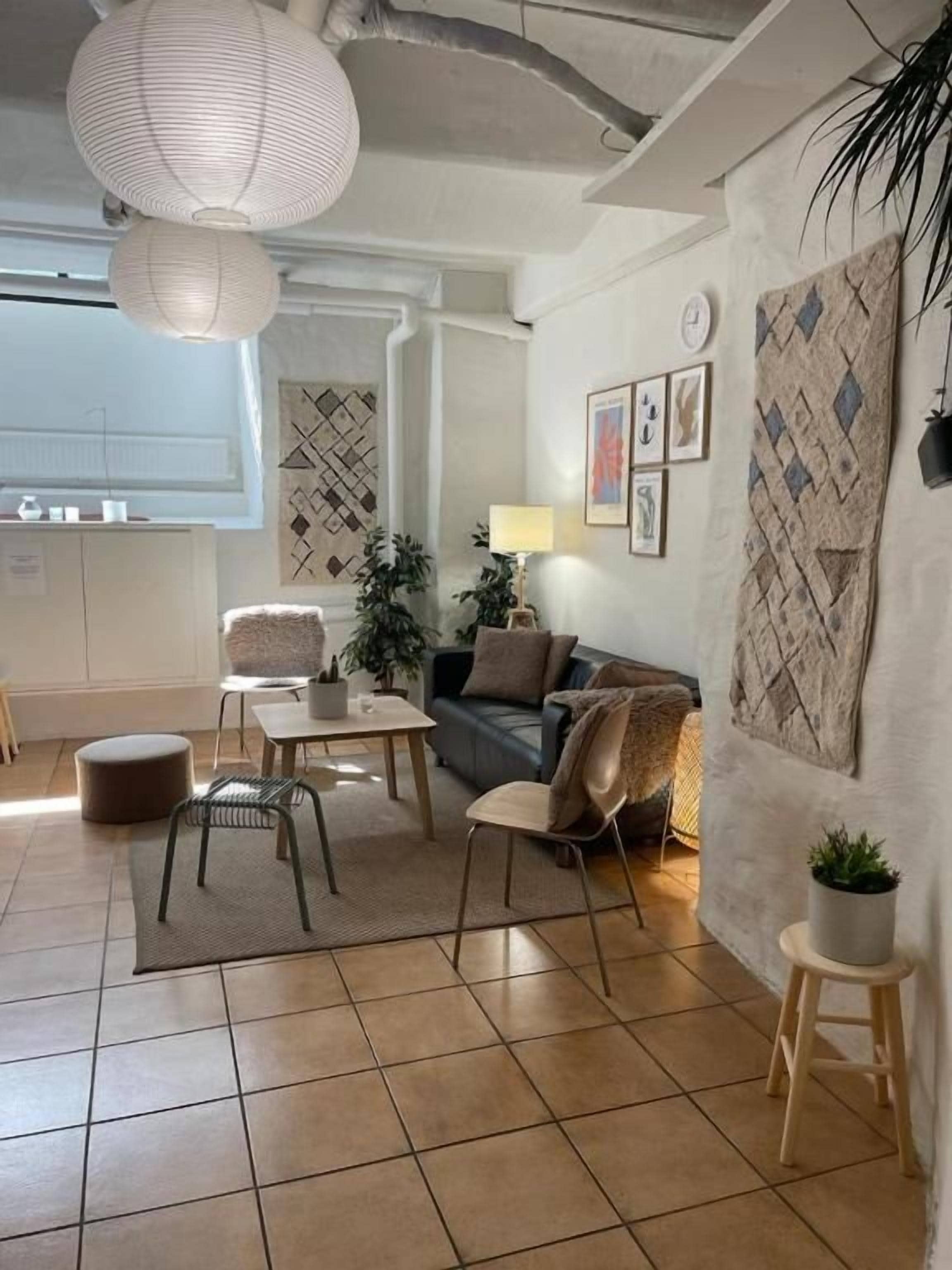 Uma sala de estar moderna com sofá, mesa de centro e cadeiras. Há uma mesa de jantar com cadeiras, um abajur e um vaso de planta. O cômodo possui piso de azulejos e uma janela grande.