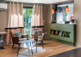 Uma área de recepção moderna com balcão verde, duas poltronas, uma pequena mesa e vista para o verde pelas janelas.