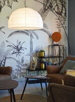 Uma sala de estar com um grande painel de folhagem tropical, uma luminária moderna, uma estante de livros e dois sofás.