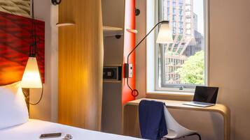 Quarto de hotel com uma cama, uma escrivaninha com um laptop, uma cadeira, um objeto cilíndrico vermelho e uma janela com vista para edifícios.