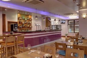 Uma área de bar com balcão roxo, diversas torneiras e uma variedade de garrafas. Mesas com cadeiras e copos de vinho estão dispostas para refeições.