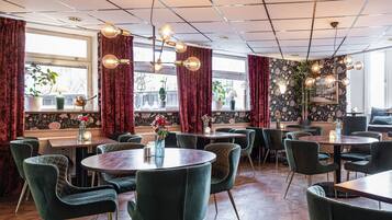 Restaurante com mesas redondas e cadeiras estofadas verdes, apresentando teto azulejado e cortinas vermelhas.