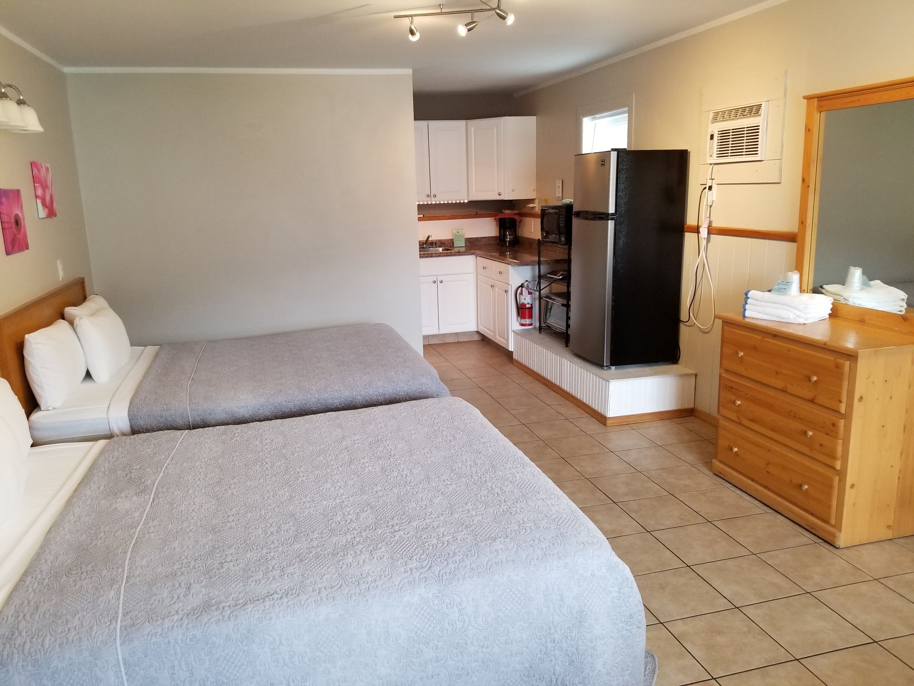 Quarto de hotel com duas camas, uma cozinha compacta, geladeira, cômoda de madeira e espelho.