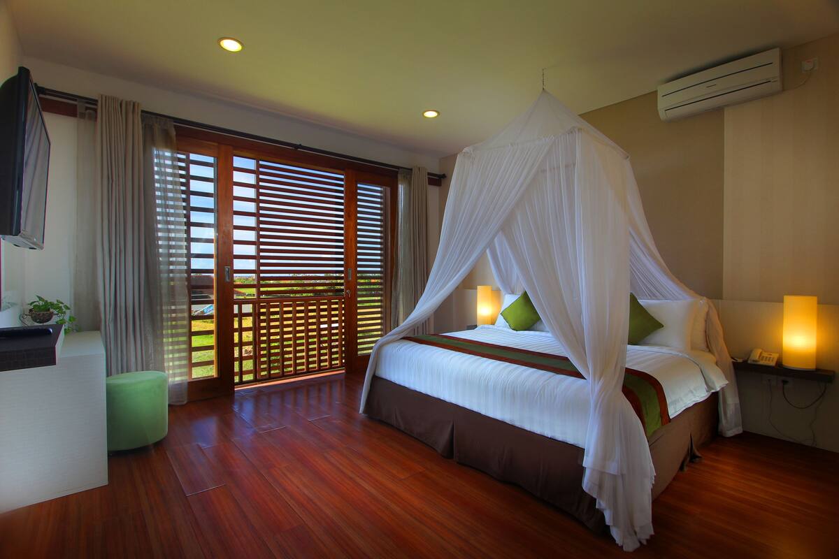 Quarto com cama de dossel, um banquinho verde, mesa de cabeceira com abajur e vista através de uma porta deslizante.
