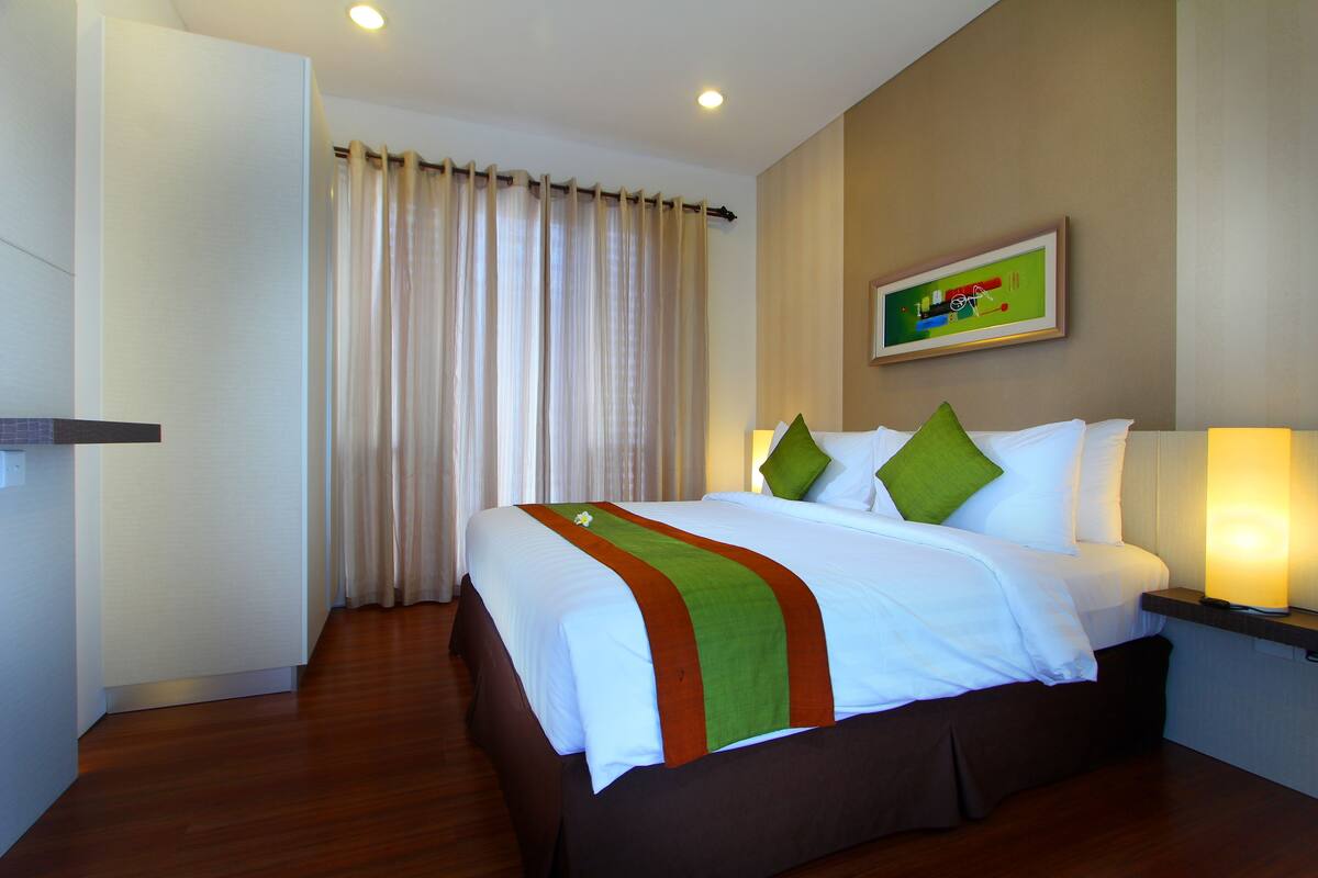 Quarto de hotel com uma cama grande, piso de madeira, cortina, um quadro na parede e mesa de cabeceira com abajur.