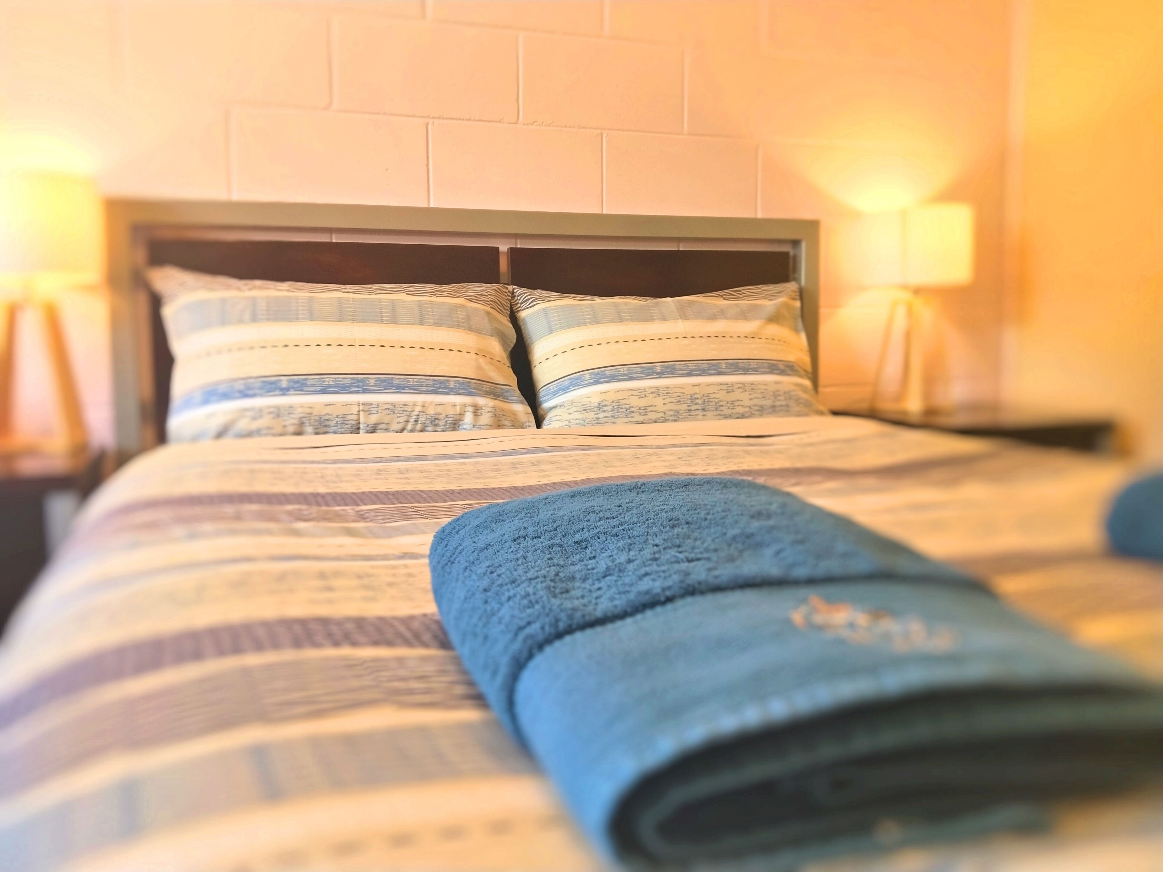 Cama bem arrumada com roupa de cama listrada e duas almofadas, um cobertor azul e uma mesa de cabeceira com abajur.