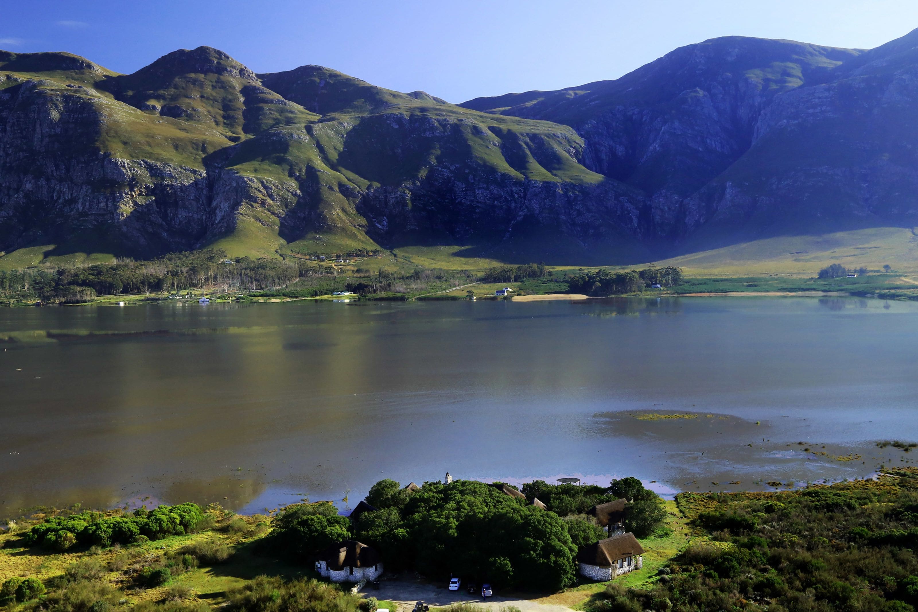 Um lago rodeado por montanhas, com um pequeno povoado e edifícios com telhados de palha perto da margem.