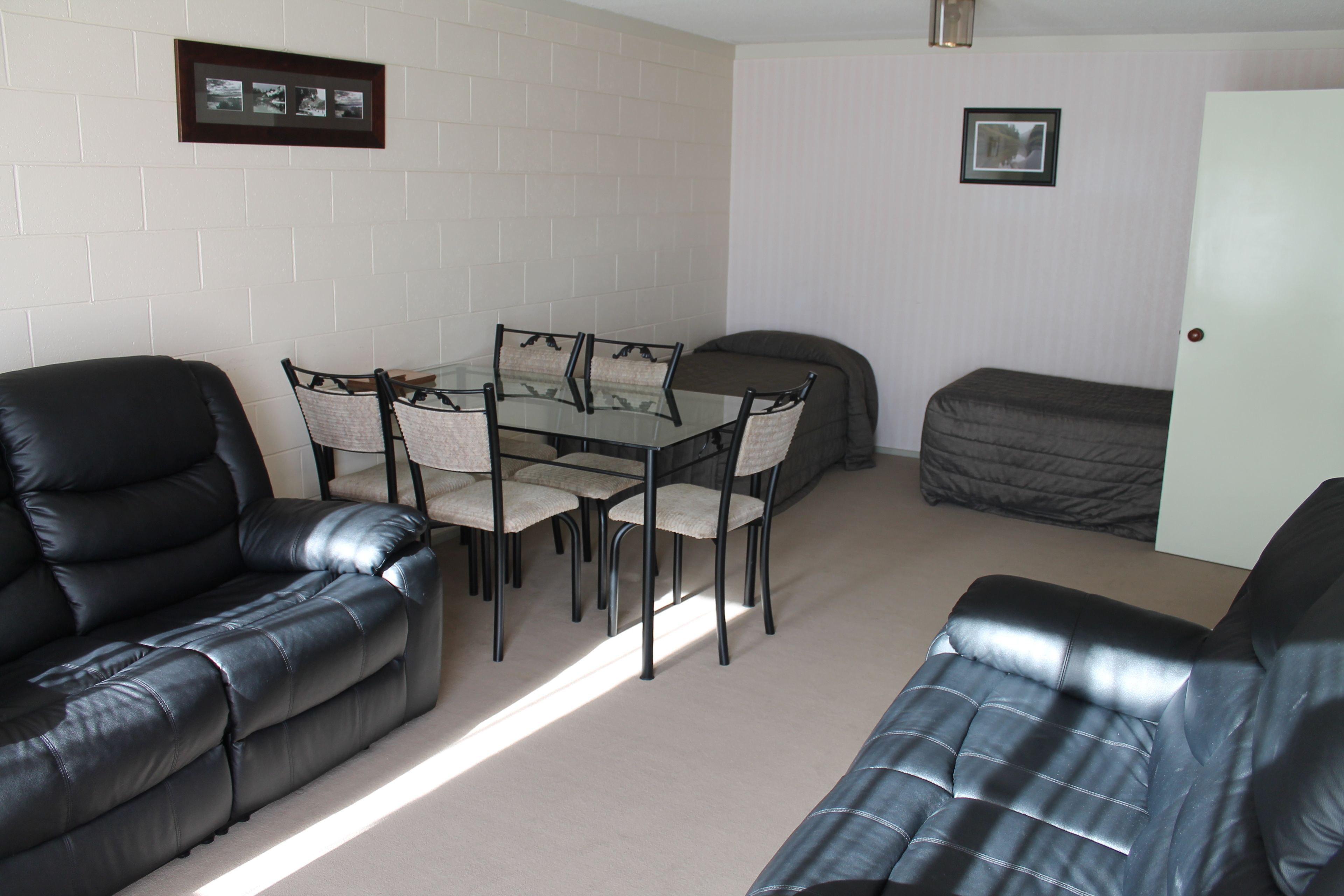 Quarto com sofá de couro preto, mesa de jantar com bancada de vidro e cadeiras, e um sofá cinzento. Há quadros nas paredes.