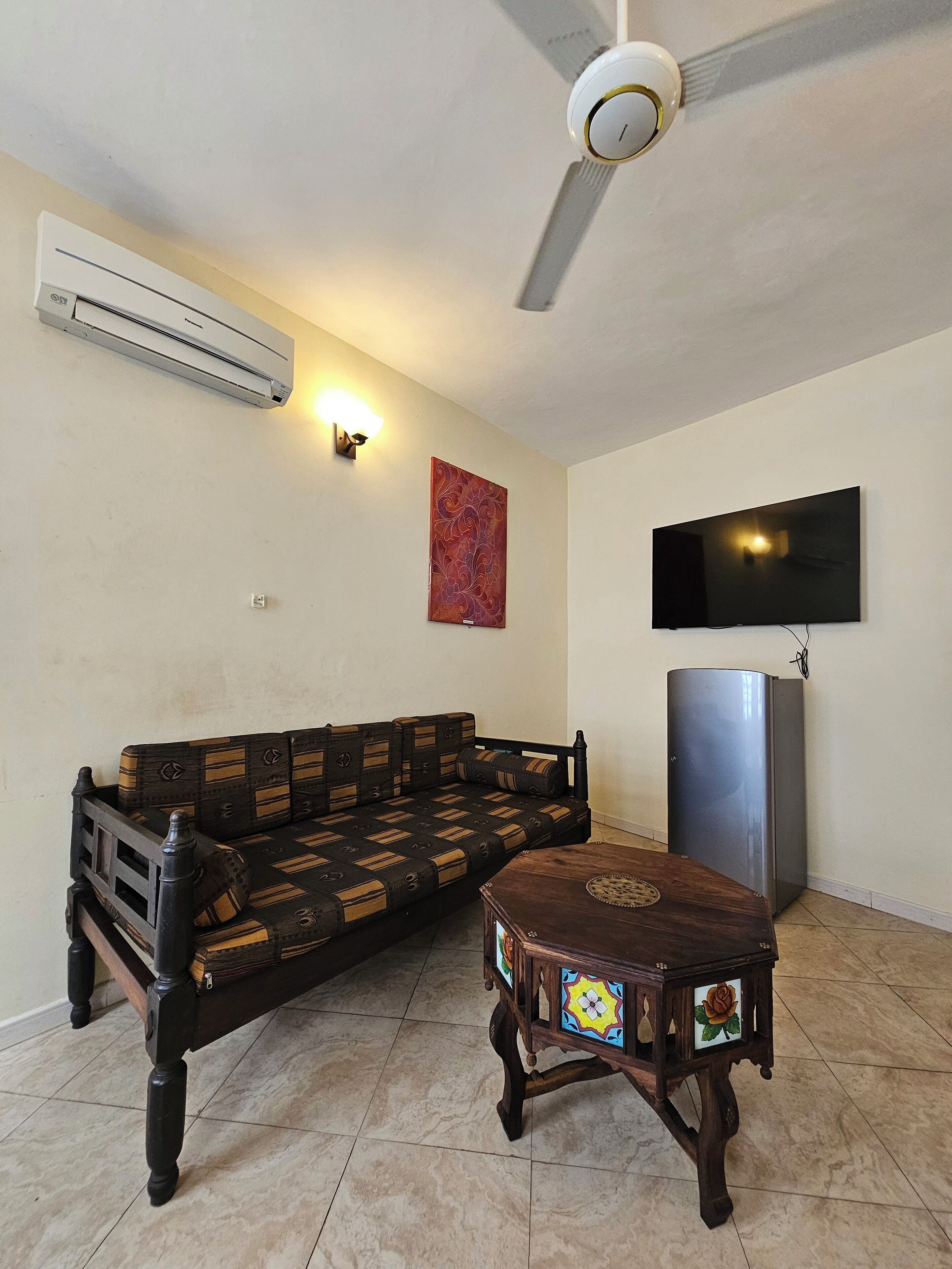 Uma sala de estar com sofá, mesa de centro, ar condicionado, ventilador de teto, TV fixada na parede e um objeto decorativo.