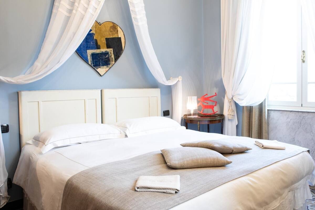Uma cama bem arrumada com lençóis brancos, um objeto decorativo na parede e uma mesa de cabeceira com abajur.