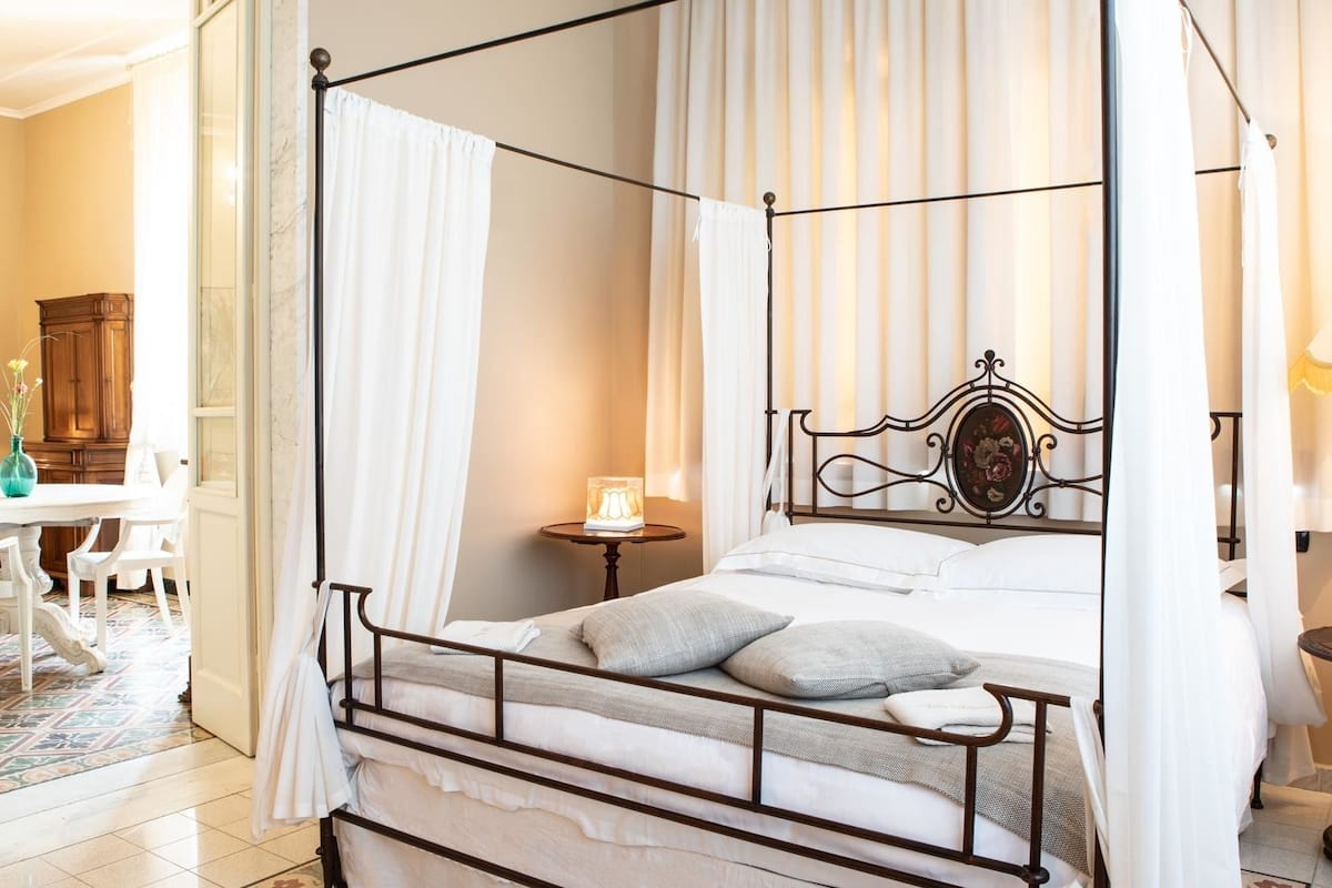 Uma cama com dossel e colunas, com cabeceira de madeira, roupa de cama branca e uma pequena mesa de cabeceira com abajur.