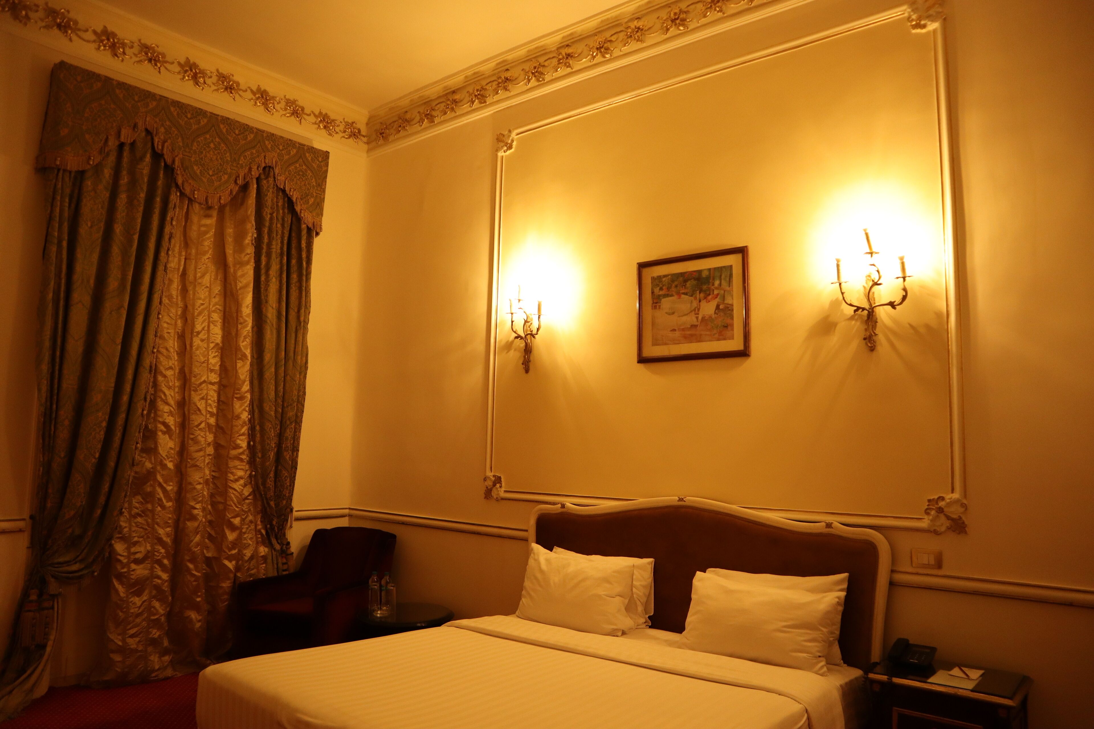 Quarto de hotel com cama, mesa de cabeceira, cadeira e luminária fixada na parede.