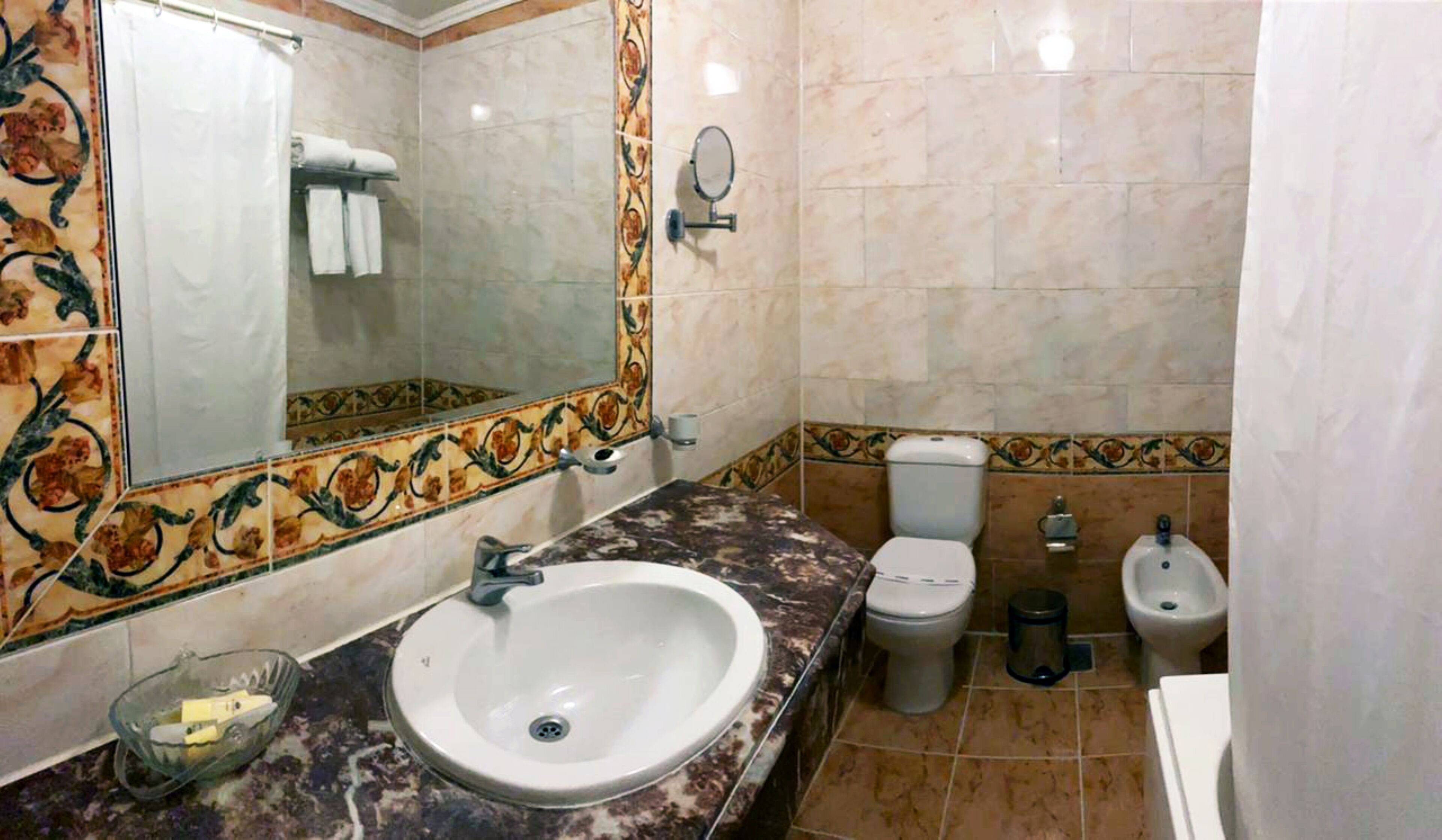Uma casa de banho com lavatório de mármore, sanita, bidé e espelho com moldura de azulejos decorativos.