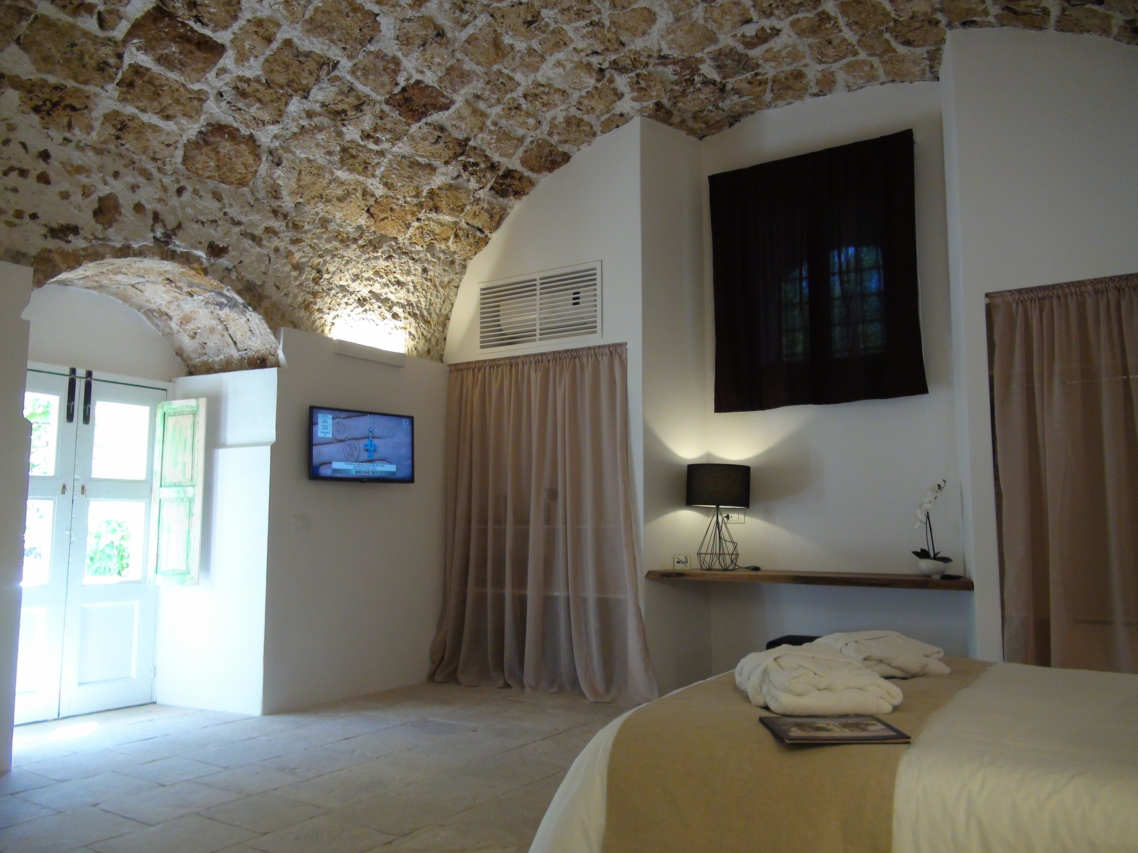 Quarto com teto de pedra, televisão, cama com edredom branco e janela com cortinas.
