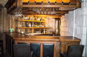 Uma área de bar com prateleiras de madeira repletas de garrafas, copos pendurados acima e banquetas estofadas.