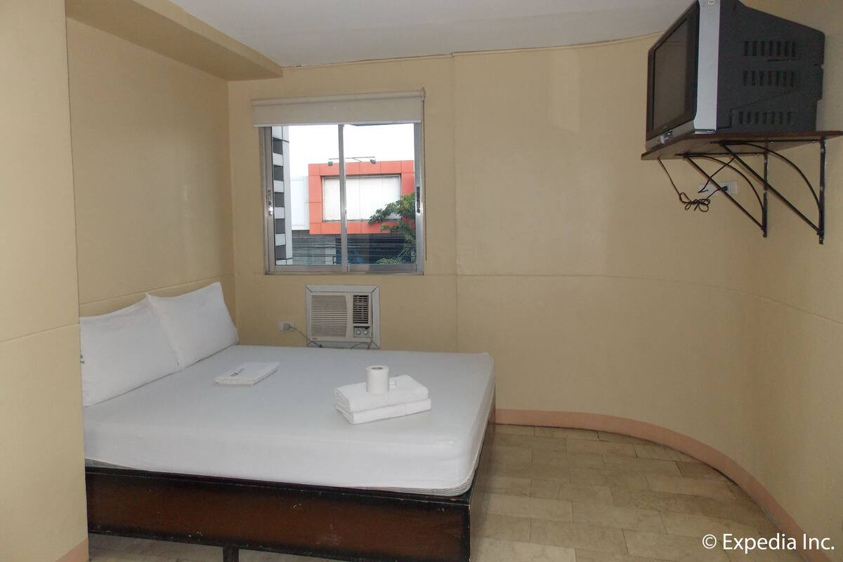Quarto de hotel pequeno com uma cama de solteiro, janela, TV fixada na parede e um pequeno aparelho de ar condicionado.