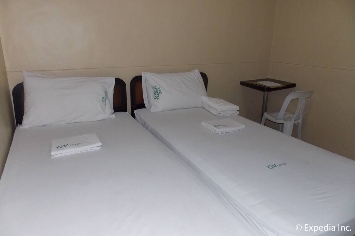 Quarto de hotel com duas camas de solteiro, cada uma com colcha e almofadas brancas, e uma mesa pequena com uma cadeira.