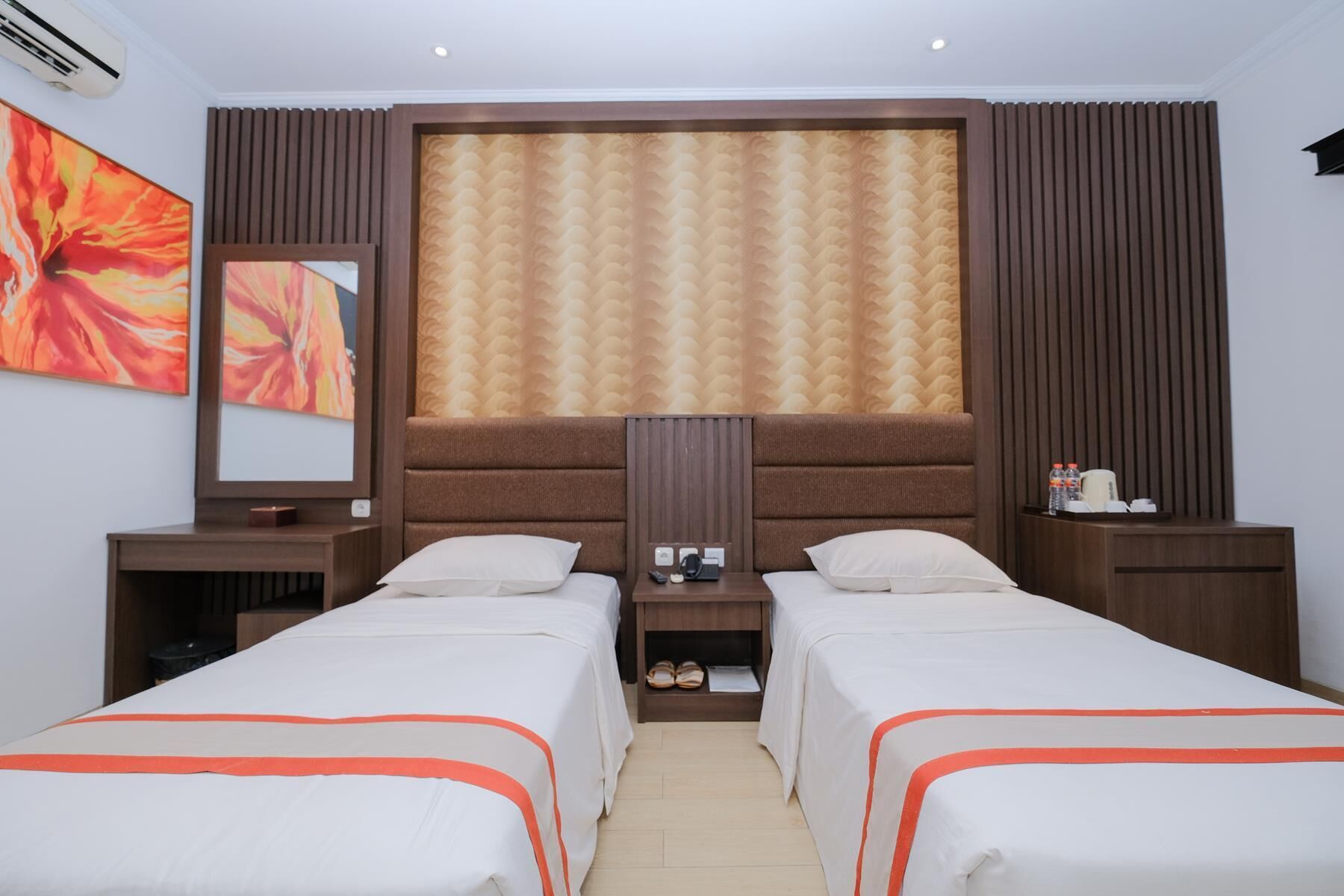 Quarto de hotel com duas camas, uma janela grande com cortinas, mesa de cabeceira com abajur e ar condicionado fixado na parede.