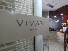 Uma área de recepção com uma placa indicando "VIVAR Hotel" e um muro de tijolos ao fundo.
