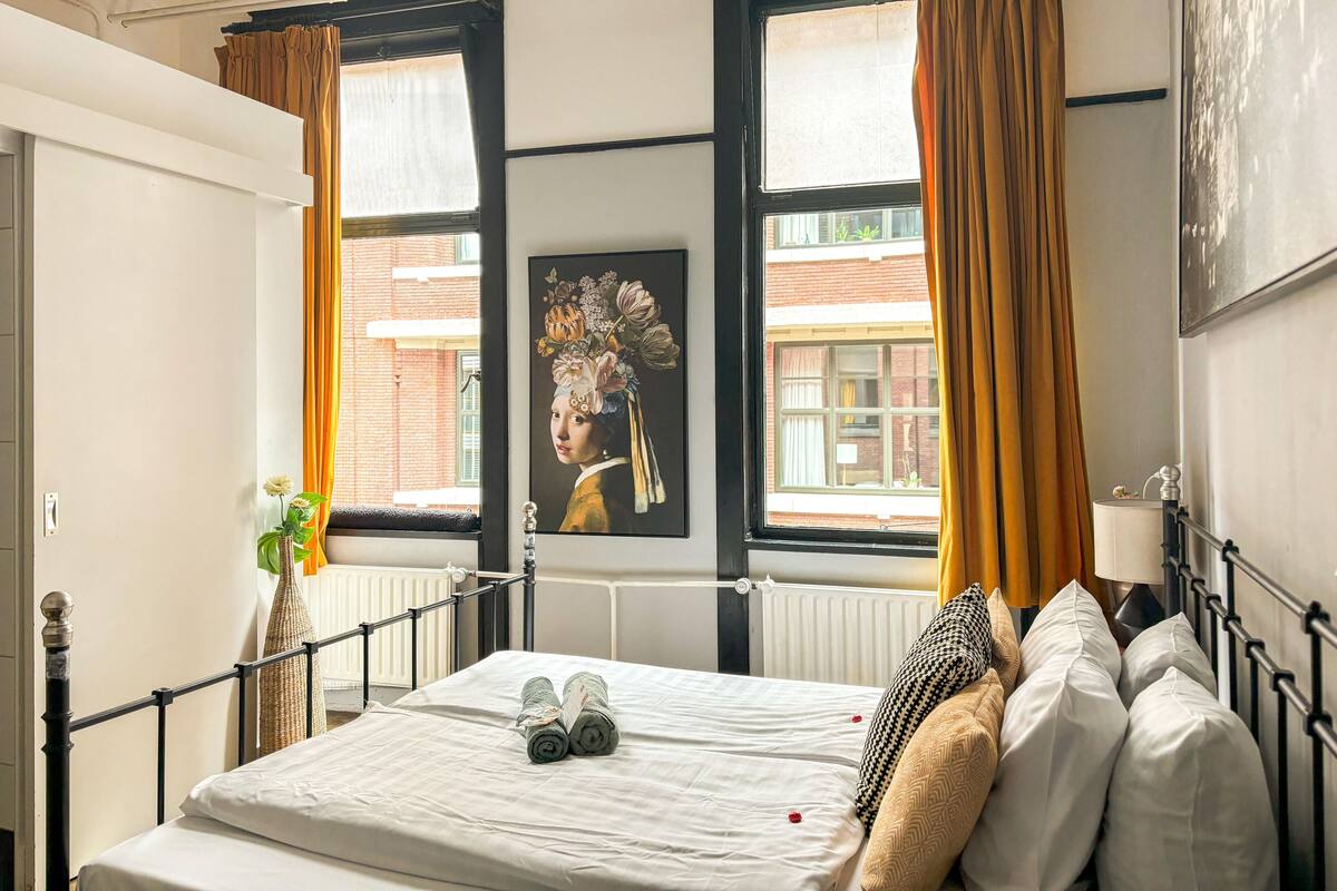 Um quarto com cama, radiador, janela com cortinas, um quadro e um vaso com flores.