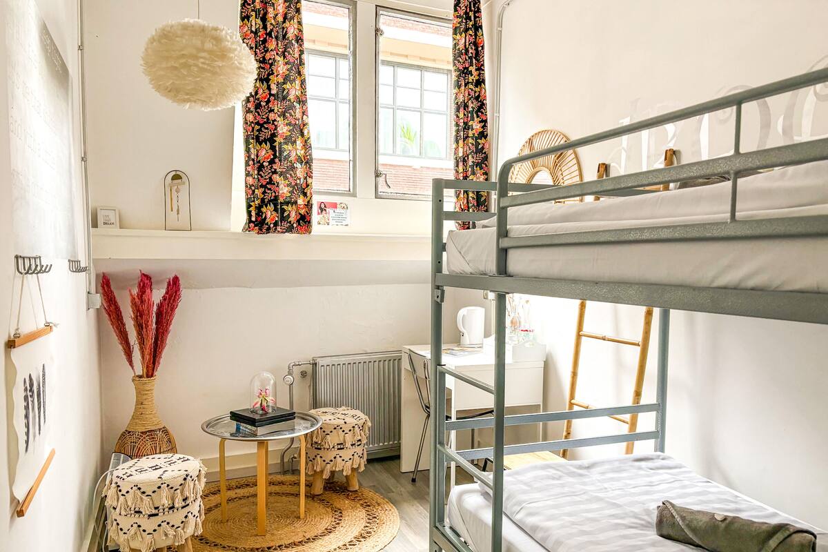 Uma cama beliche com colchão branco, uma mesinha redonda com um tapete tecido, uma prateleira na parede com objetos decorativos e uma janela com cortinas florais.