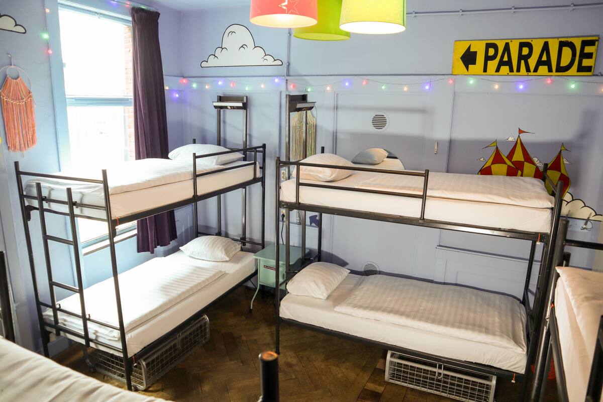 Um dormitório com beliches, um letreiro 'PARADE', luzes de cordão e uma janela com cortinas.