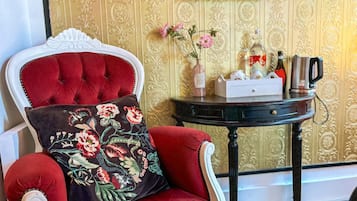 Uma poltrona vermelha estofada com almofada florida, uma mesa pequena com um vaso de flores e um quadro na parede.