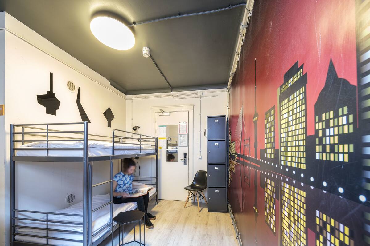 Quarto de dormitório com beliches, escrivaninha e cadeira. Um mural na parede retrata um panorama urbano.