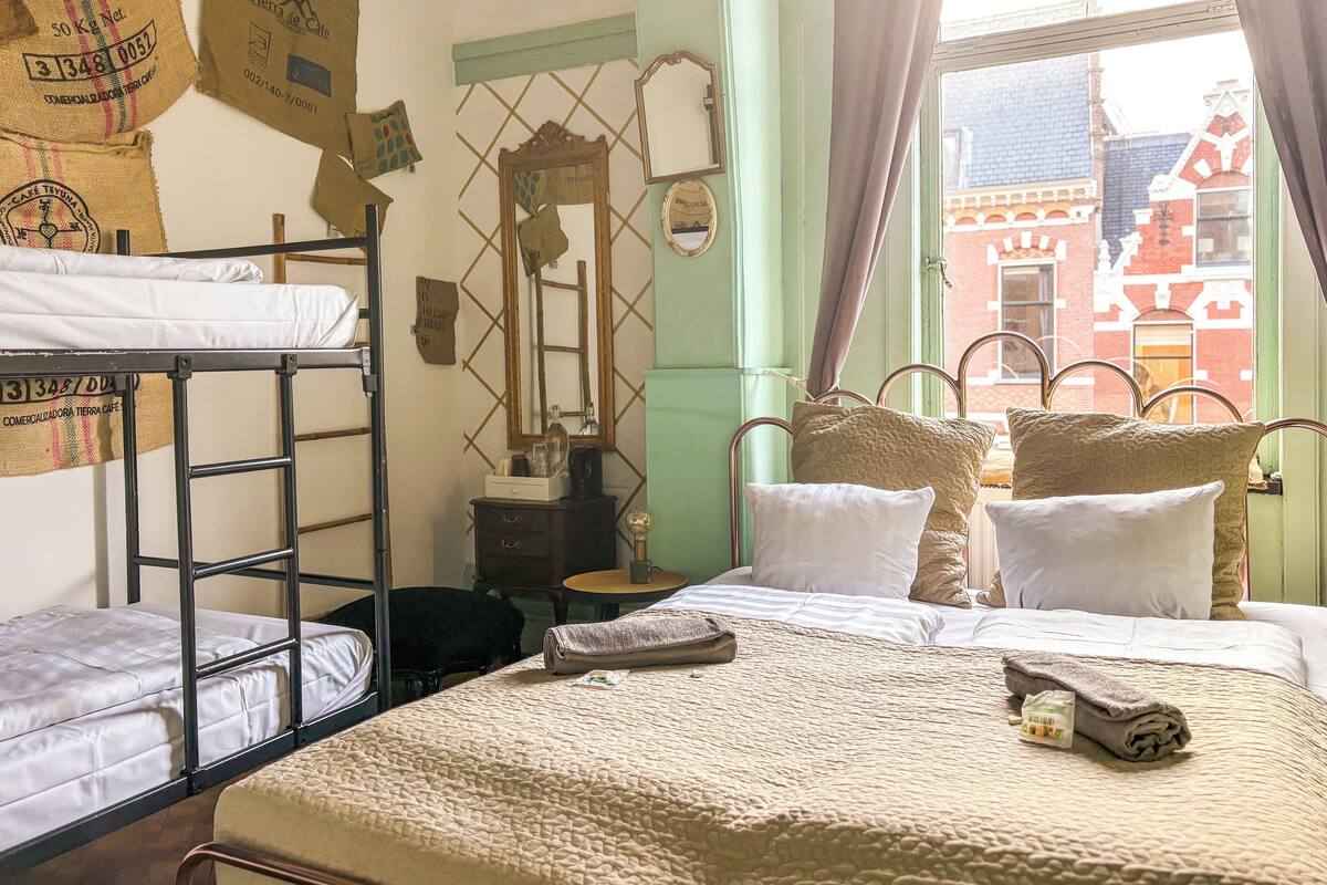 Quarto de dormitório com beliches, uma pequena mesa com abajur, espelho e janela com vista para edifícios.