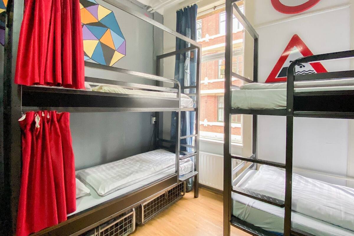 Quarto em dormitório com beliches, um ornamento geométrico na parede e vista para um edifício pela janela.