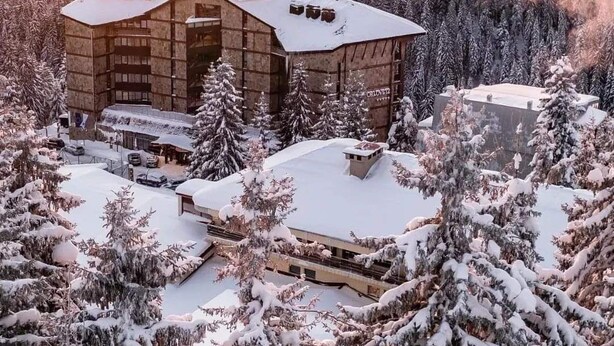Um edifício moderno com telhado plano e várias janelas, cercado por árvores cobertas de neve e um cenário montanhoso.