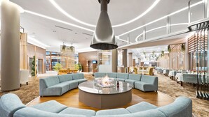 Um hall moderno com área de estar circular central, lareira e um restaurante ao fundo.