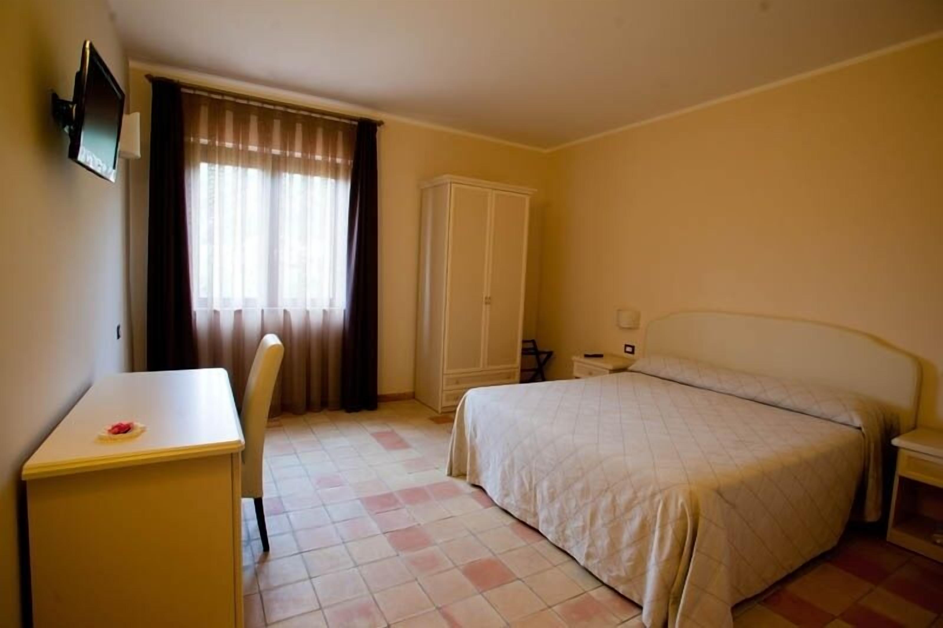 Quarto de hotel com cama, escrivaninha, cadeira, guarda-roupa e televisão na parede.