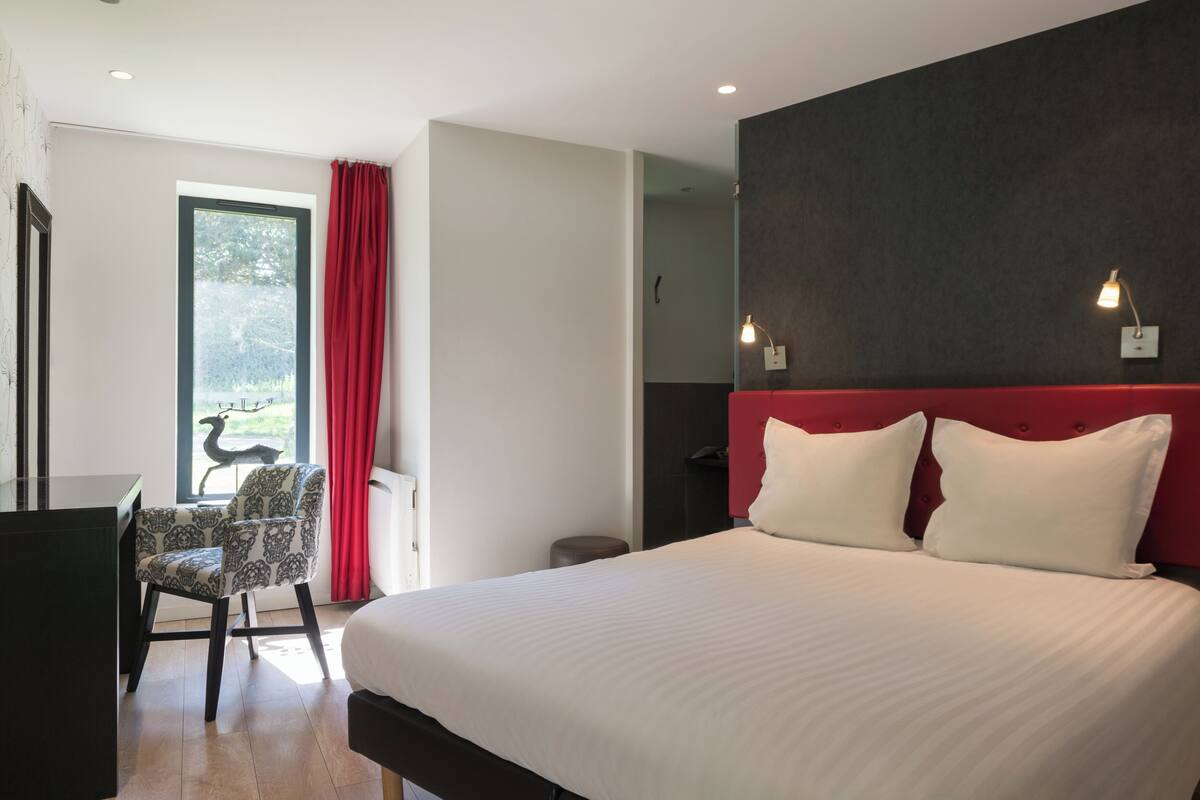 Quarto de hotel com uma cama grande, escrivaninha, cadeira e janela com cortinas.