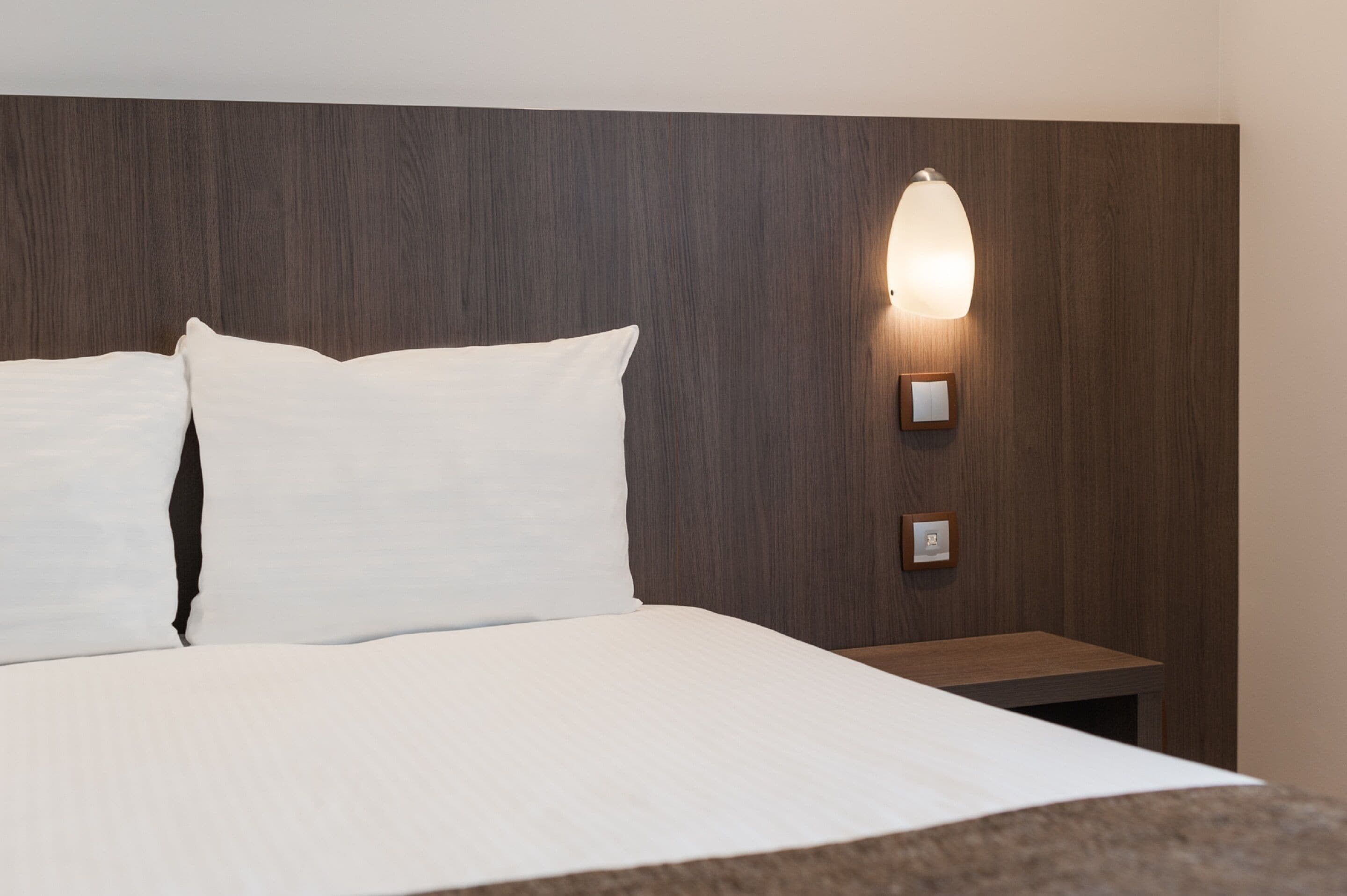 Cama bem arrumada com lençóis brancos e duas almofadas, cabeceira de madeira e luminária fixada na parede.