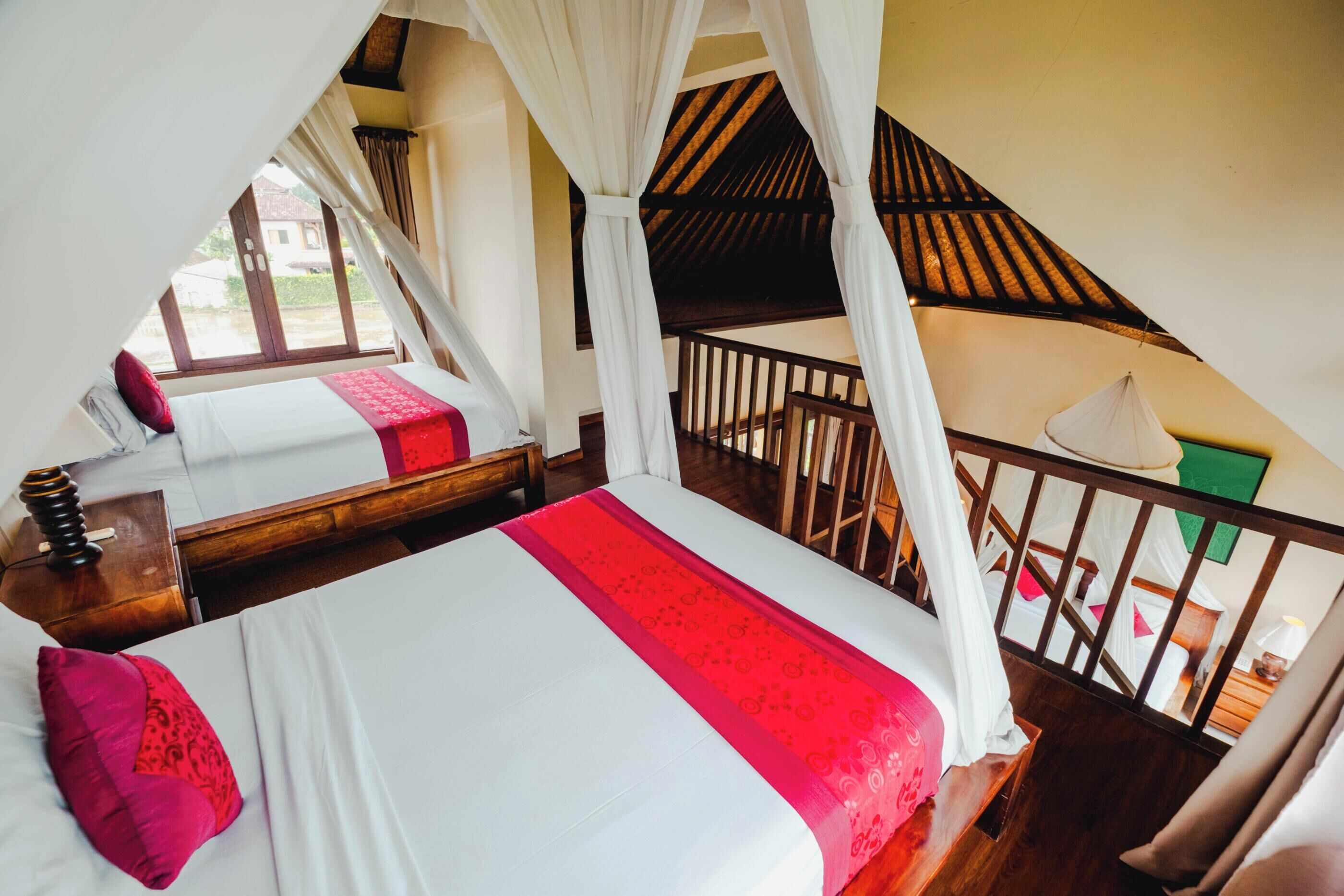 Quarto com cama, mesa de cabeceira, corrimão de madeira e vista para o exterior.