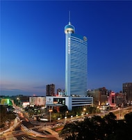 Um hotel com muitos andares e um topo cilíndrico característico, rodeado por um horizonte urbano ao entardecer.