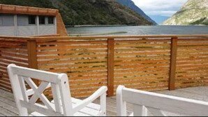Um deck de madeira com cadeiras brancas, com vista para um corpo de água e uma paisagem montanhosa.