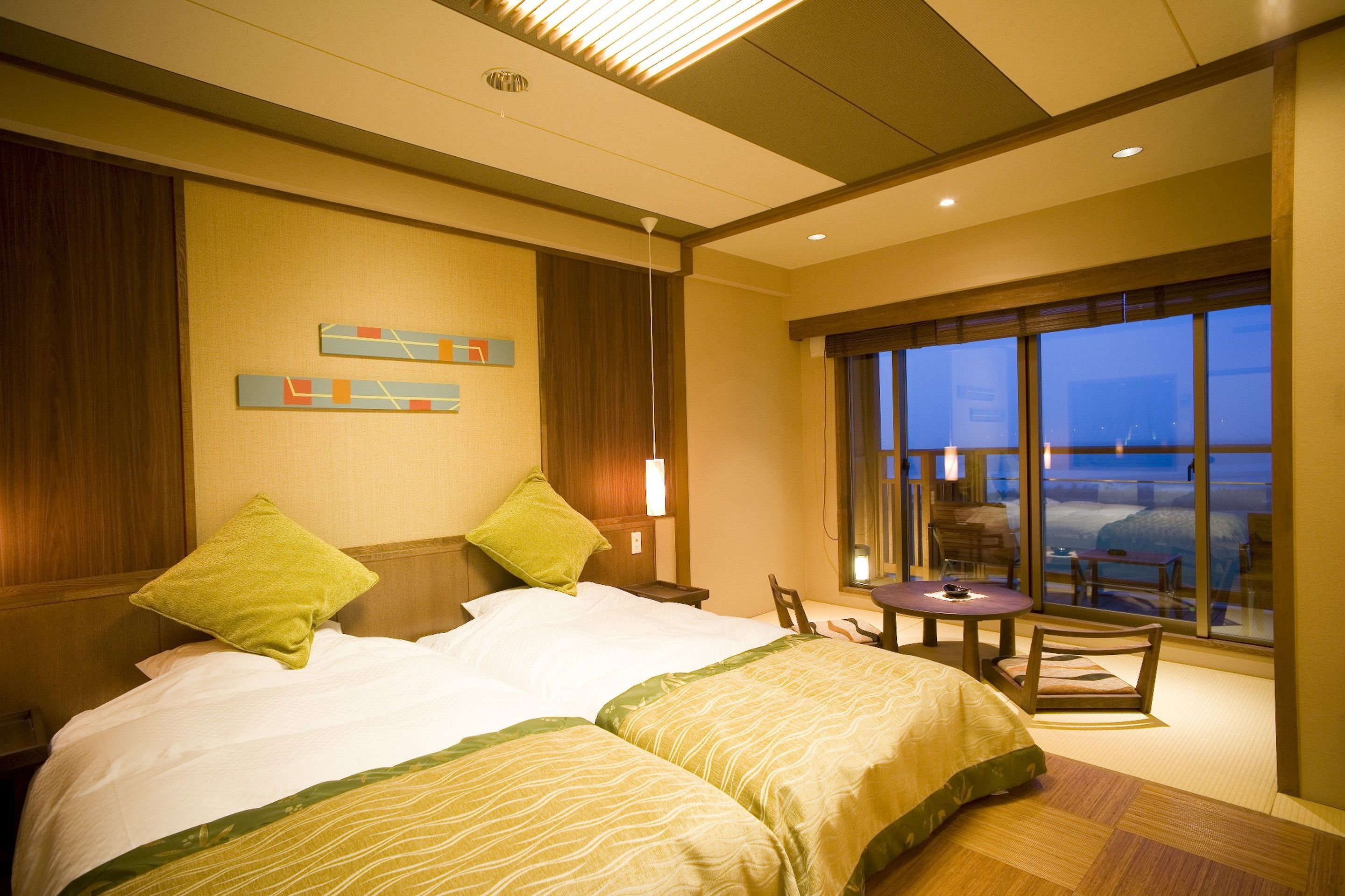 Quarto de hotel com uma cama grande, duas almofadas, uma mesa de cabeceira, uma cadeira, uma mesinha e uma janela ampla com vista.