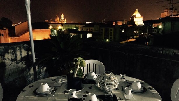 Mesa posta para refeição com pratos, xícaras e bule, com vista para a paisagem urbana à noite.