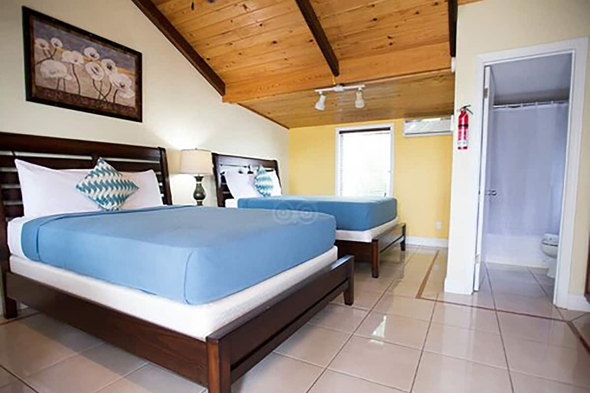 Quarto de hotel com duas camas, vigas de madeira no teto, um quadro na parede e um banheiro visível através de uma porta aberta.