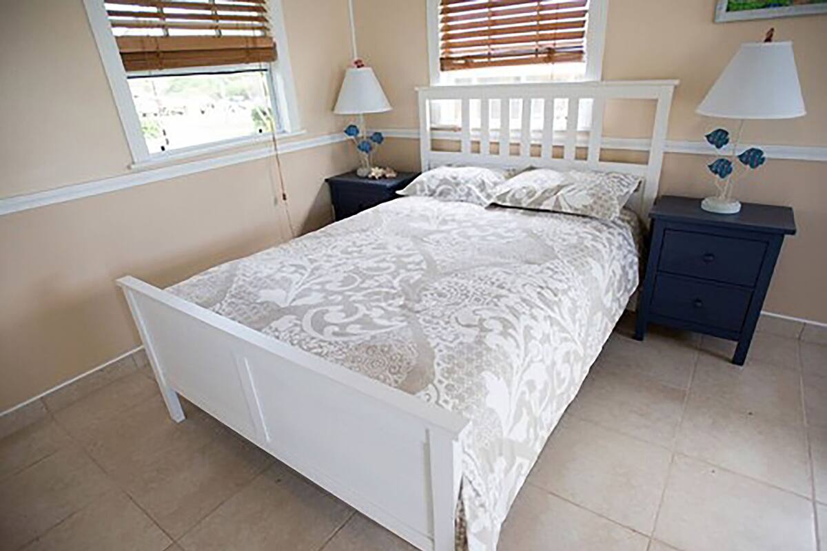 Quarto com cama branca, duas mesas de cabeceira com luminárias e uma janela com persianas.