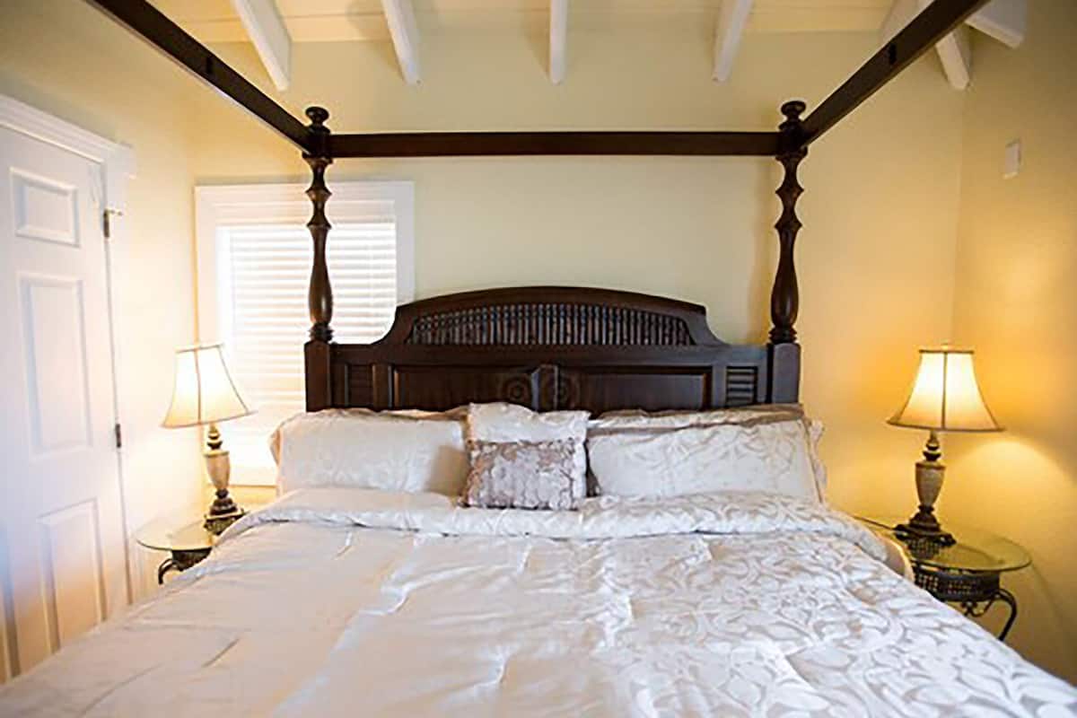 Uma cama com dossel e cabeceira de madeira, duas mesas de cabeceira com abajures e uma porta à esquerda.