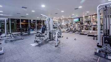 Uma academia moderna com diversos equipamentos de exercício, janelas amplas e uma tela digital.